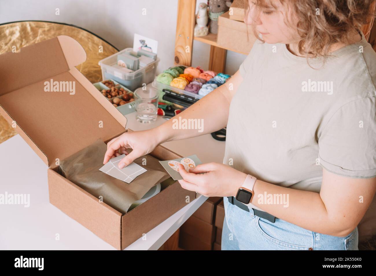 Smiling blond curly woman packing and wrapping shipment with postcards ...