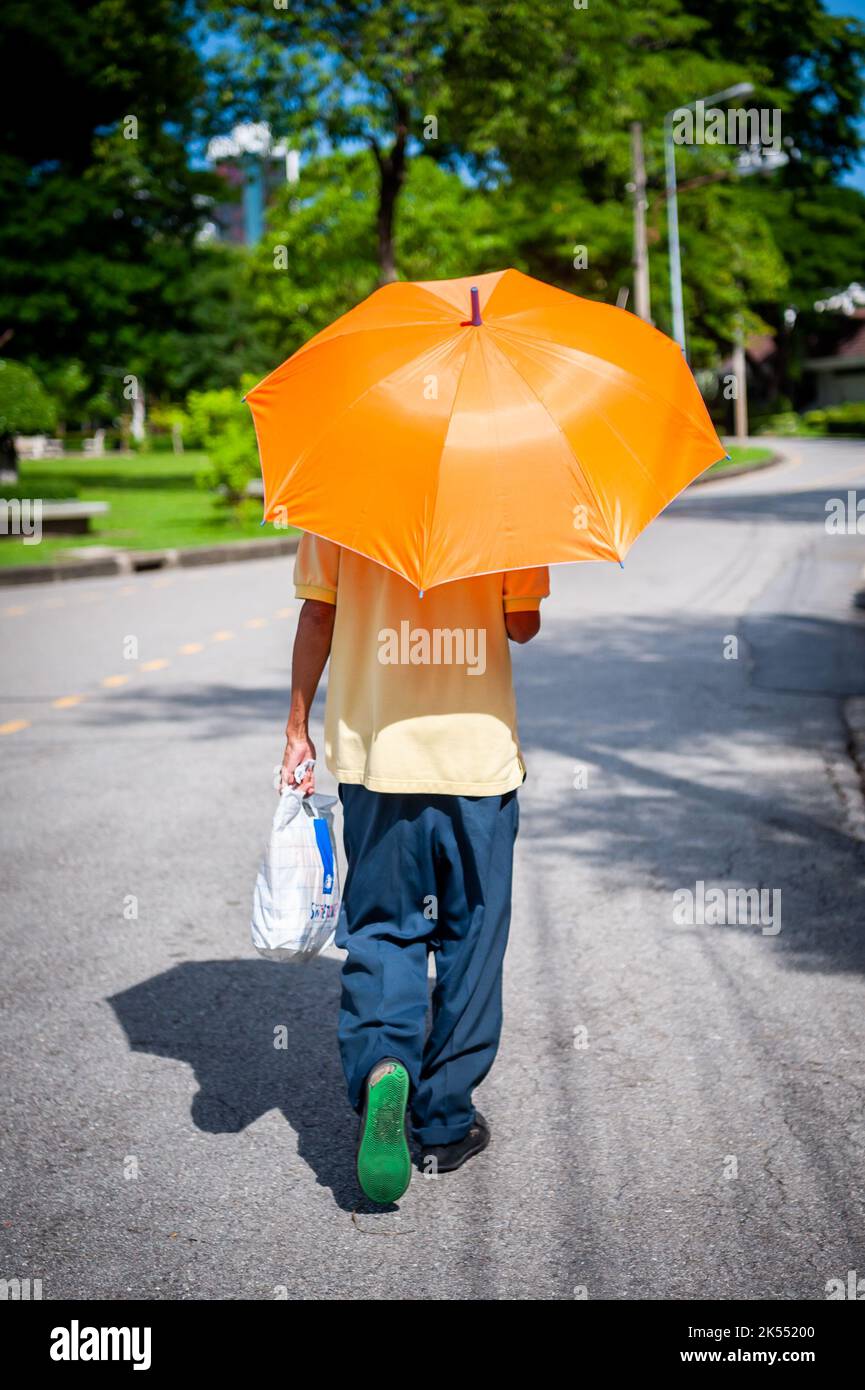 A Thai man holding an orange umbrella to protect against the hot sun ...