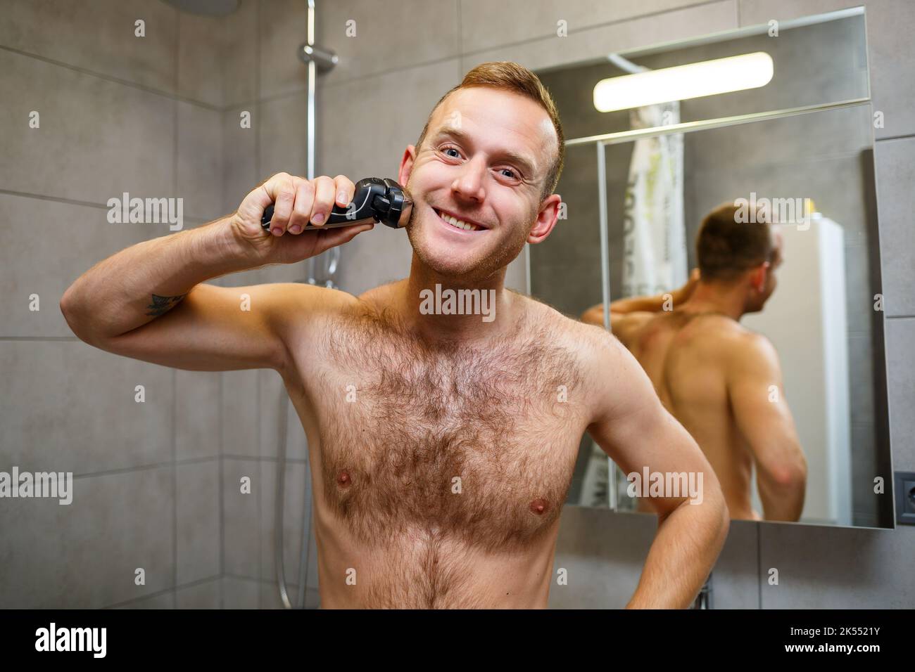 A man shaves his face with an electric razor in front of a mirror. Skin ...