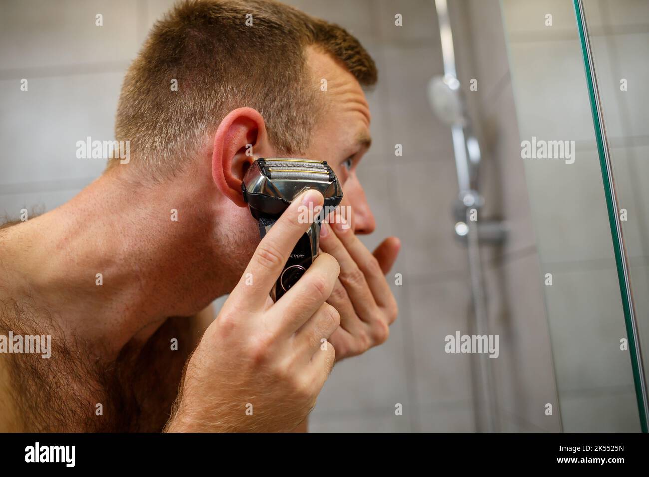 A man shaves his face with an electric razor in front of a mirror. Skin ...