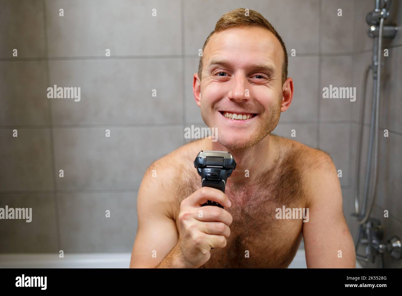 A man shaves his face with an electric razor in front of a mirror. Skin ...