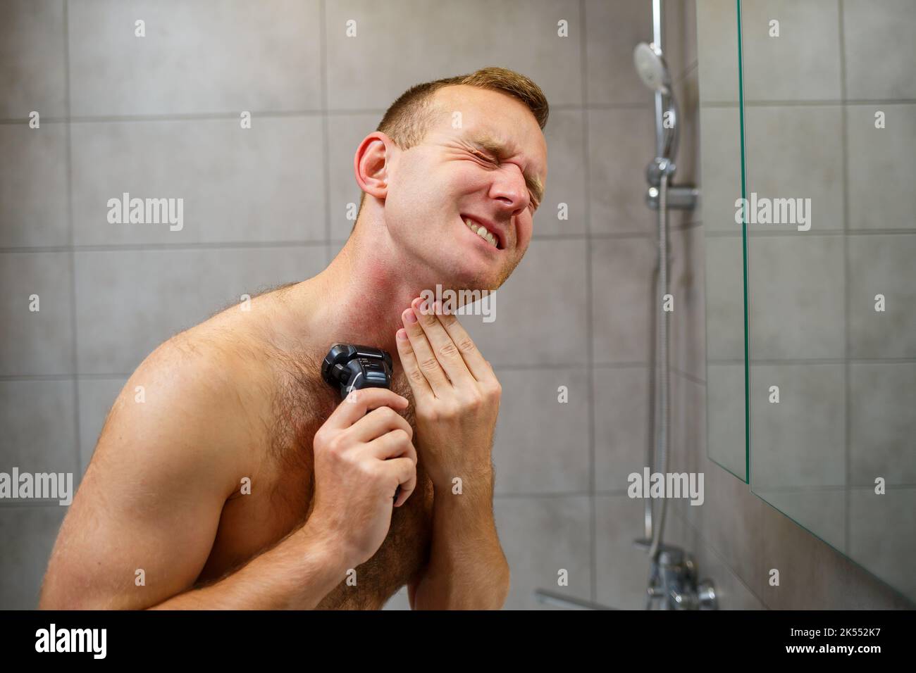 A man shaves his face with an electric razor in front of a mirror. Skin ...