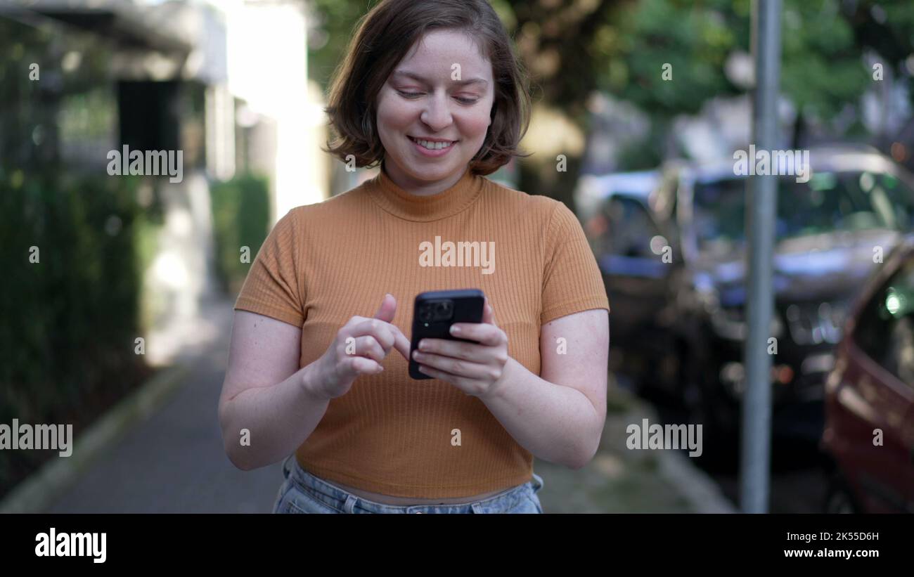 Happy young woman looking at smartphone walking in city street. Girl checking cellphone while ...