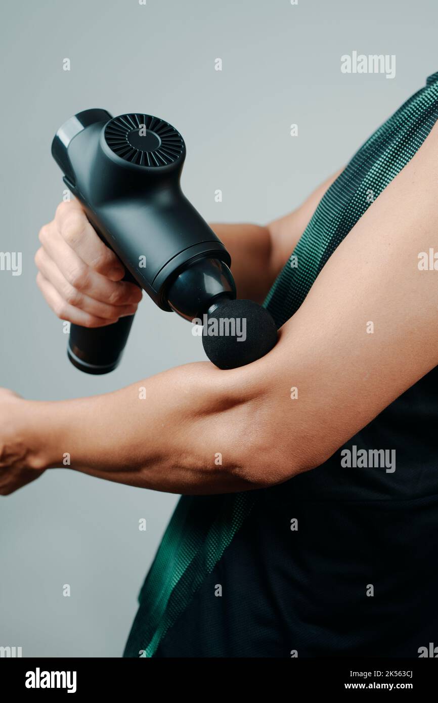 a young caucasian sportsman, wearing sports clothes, uses a massage gun ...