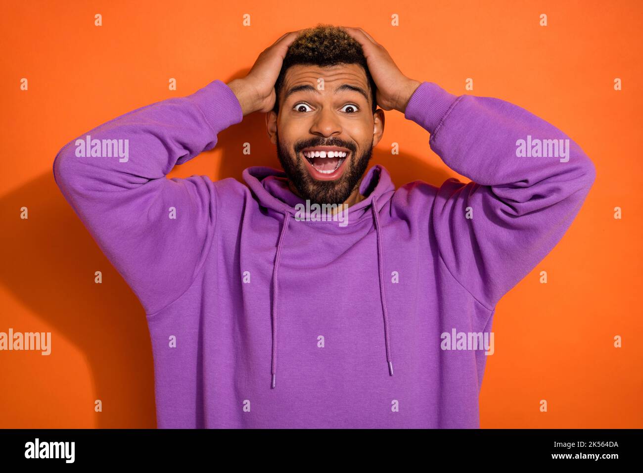 Photo of excited man wear purple hoodie raise up arms touch head ...