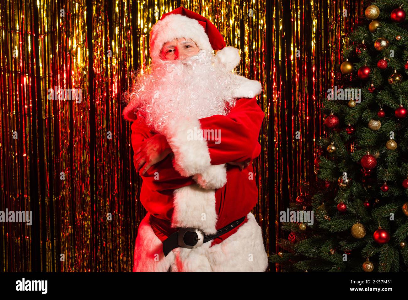 Santa claus in costume crossing arms and looking at camera near christmas tree and tinsel,stock ...