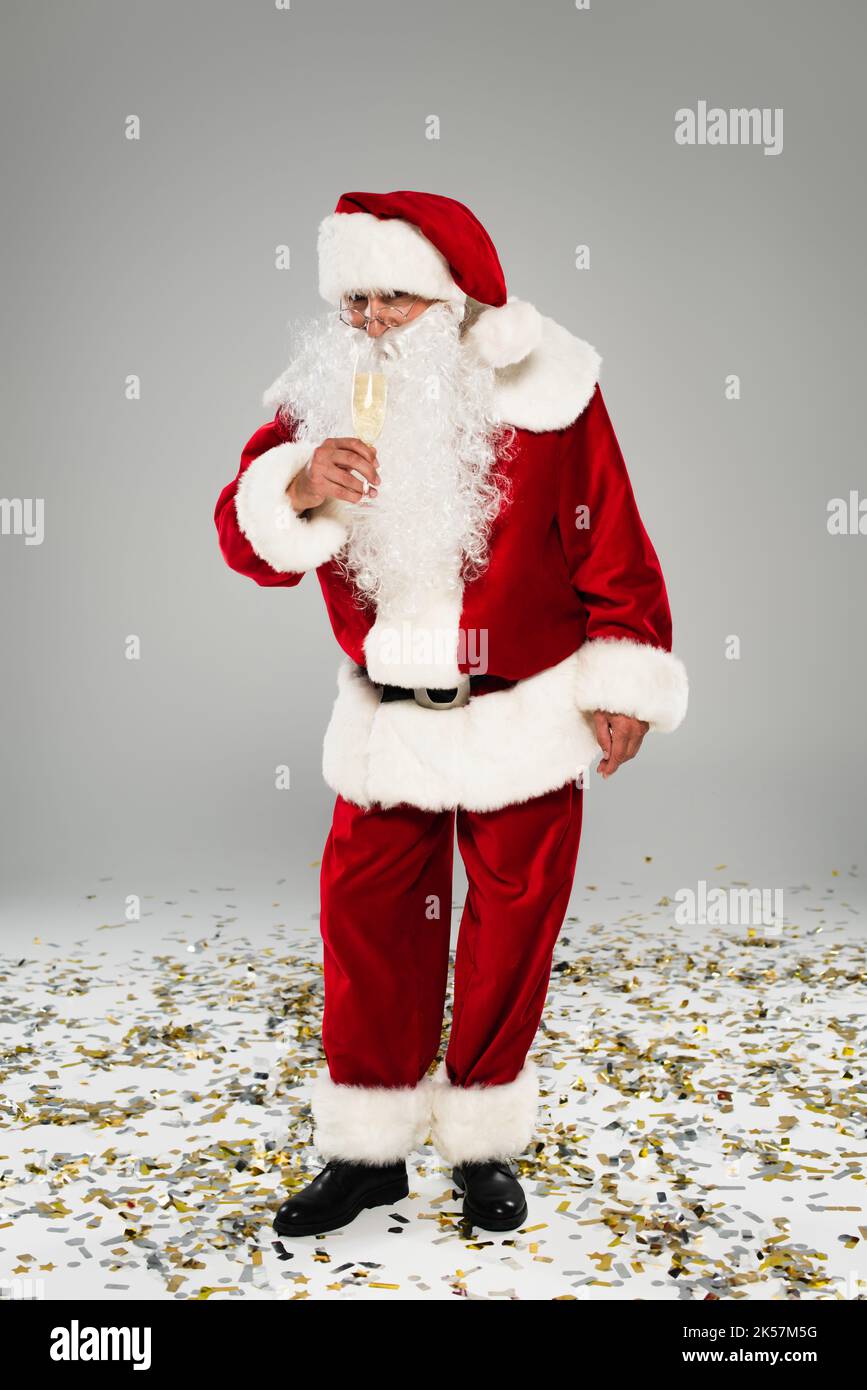 Santa claus in costume holding glass of champagne near confetti on grey ...