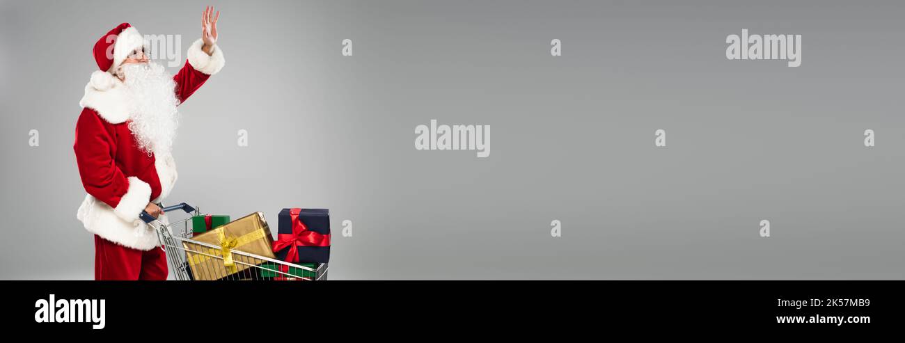 Santa claus waving hand near shopping cart with gift boxes isolated on ...