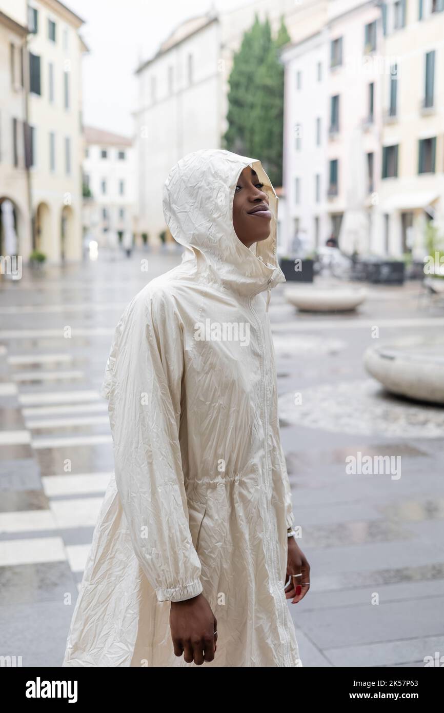 Smiling african american woman in raincoat standing on blurred urban ...