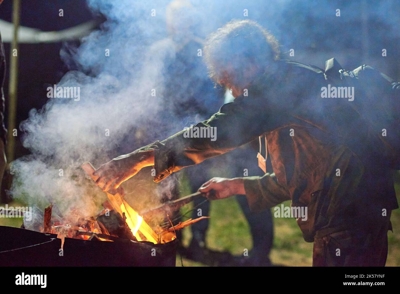 An unrecognizable man with long hair throws wood on a fire lit in a ...