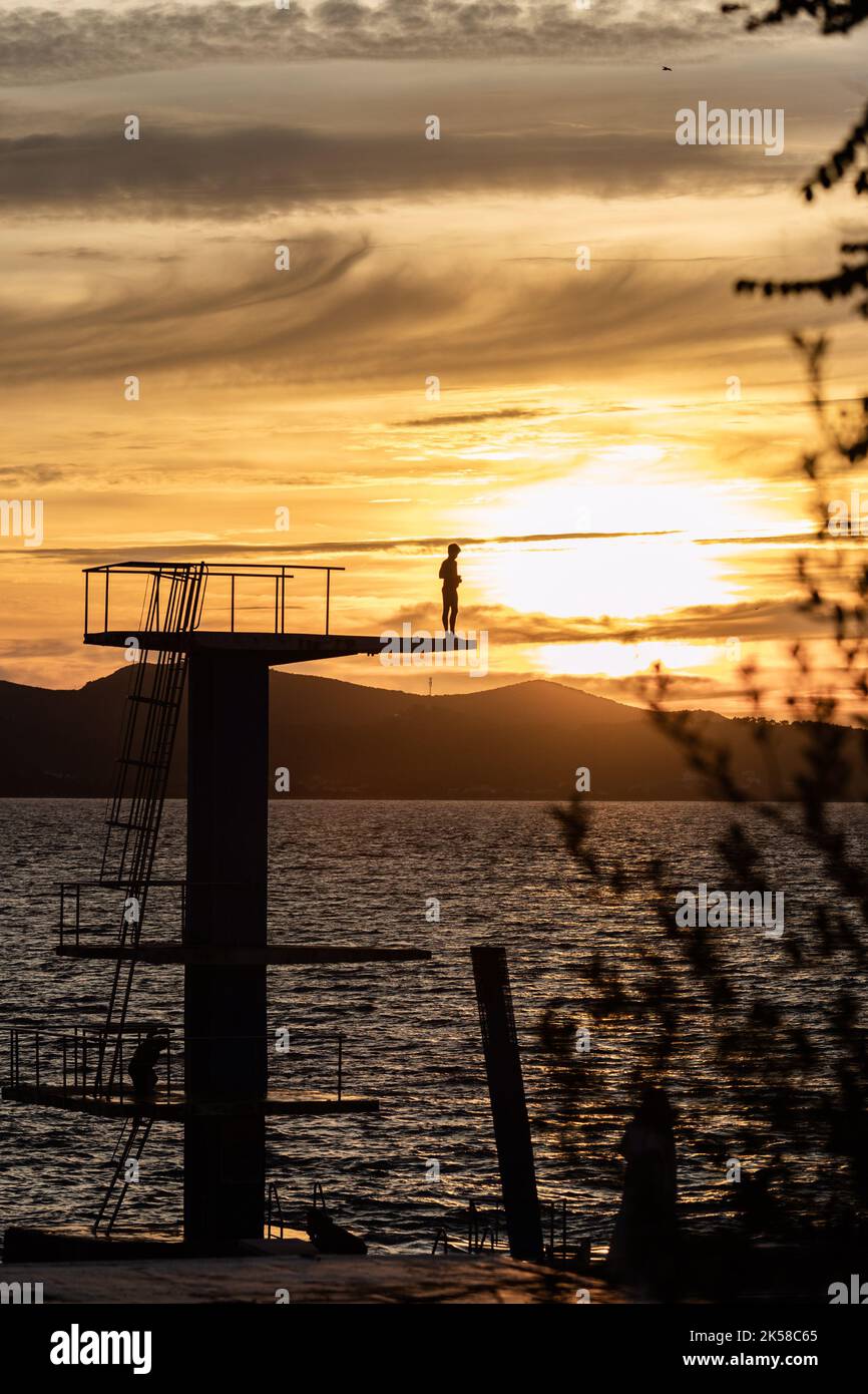 Boy jumps off the diving platform into the Adriatic Sea during sunset ...