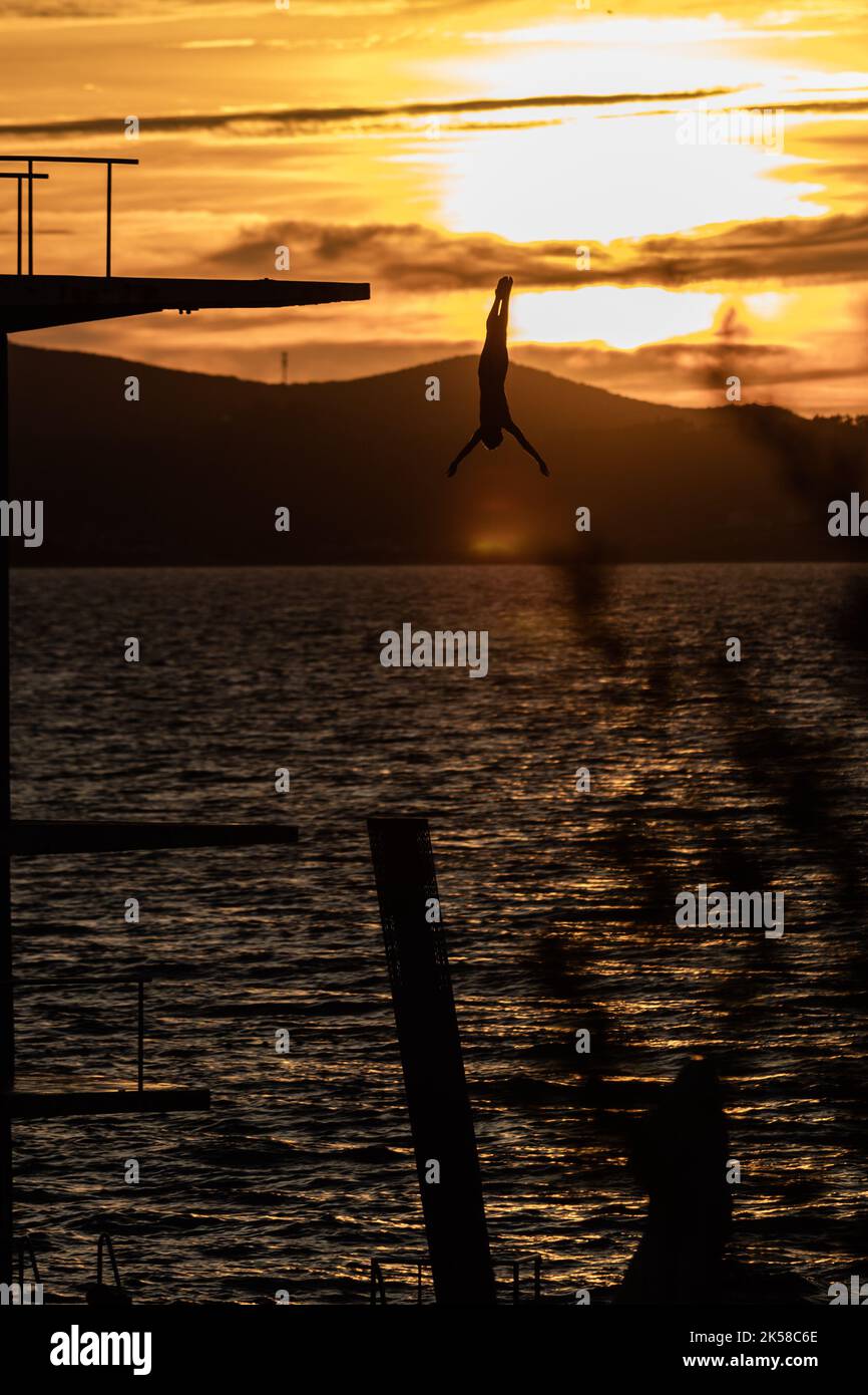 Boy jumps off the diving platform into the Adriatic Sea during sunset ...