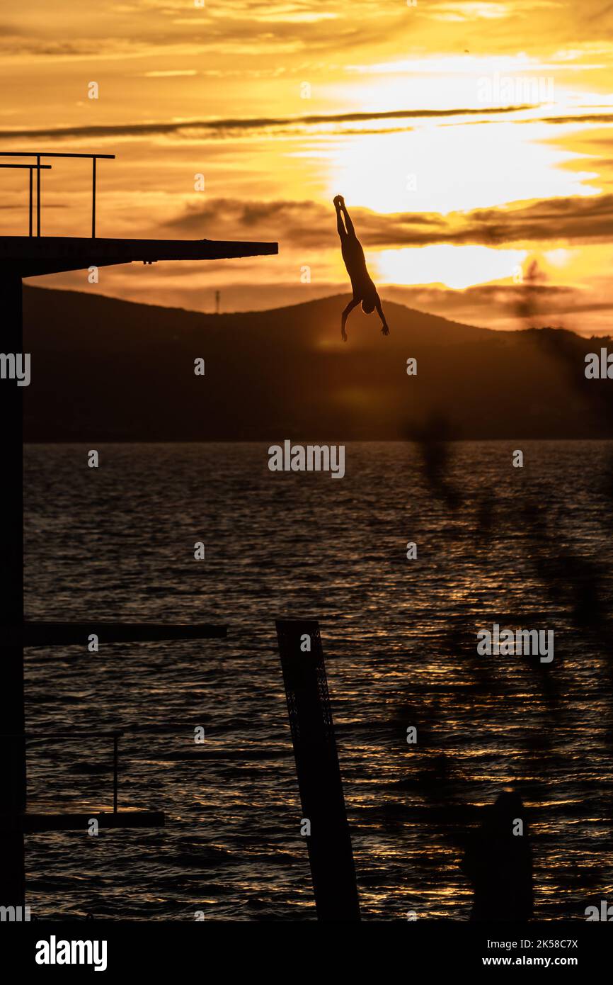 Boy jumps off the diving platform into the Adriatic Sea during sunset ...