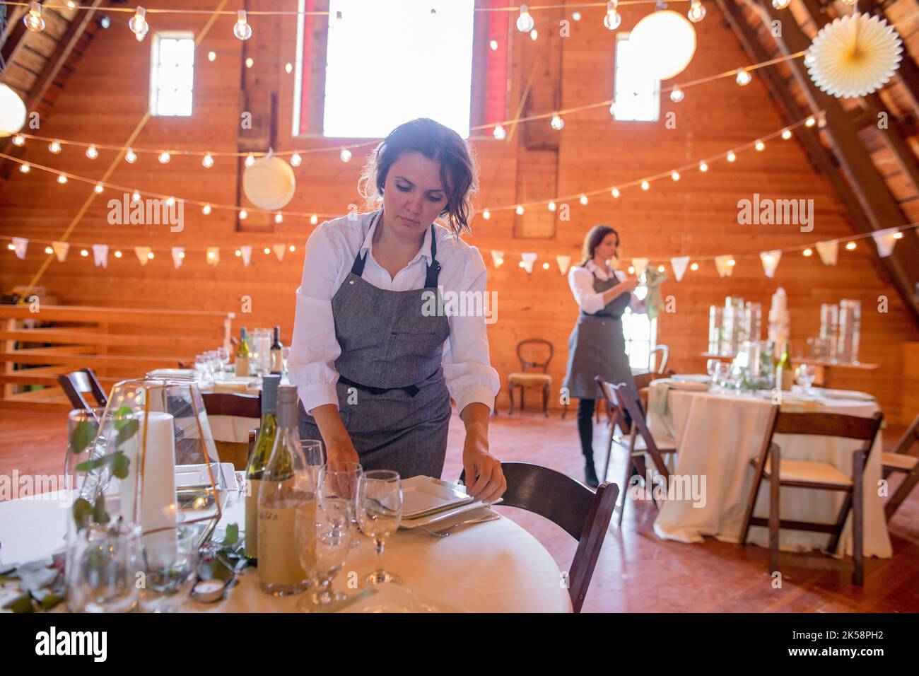 Female Server Preparing Table For Wedding Reception In Barn Stock Photo female-server-preparing-table-for-wedding-reception-in-barn-stock-photo