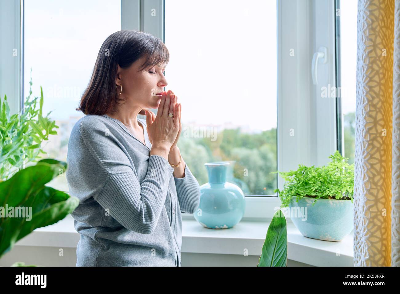 Middle-aged serious woman praying near window at home Stock Photo - Alamy