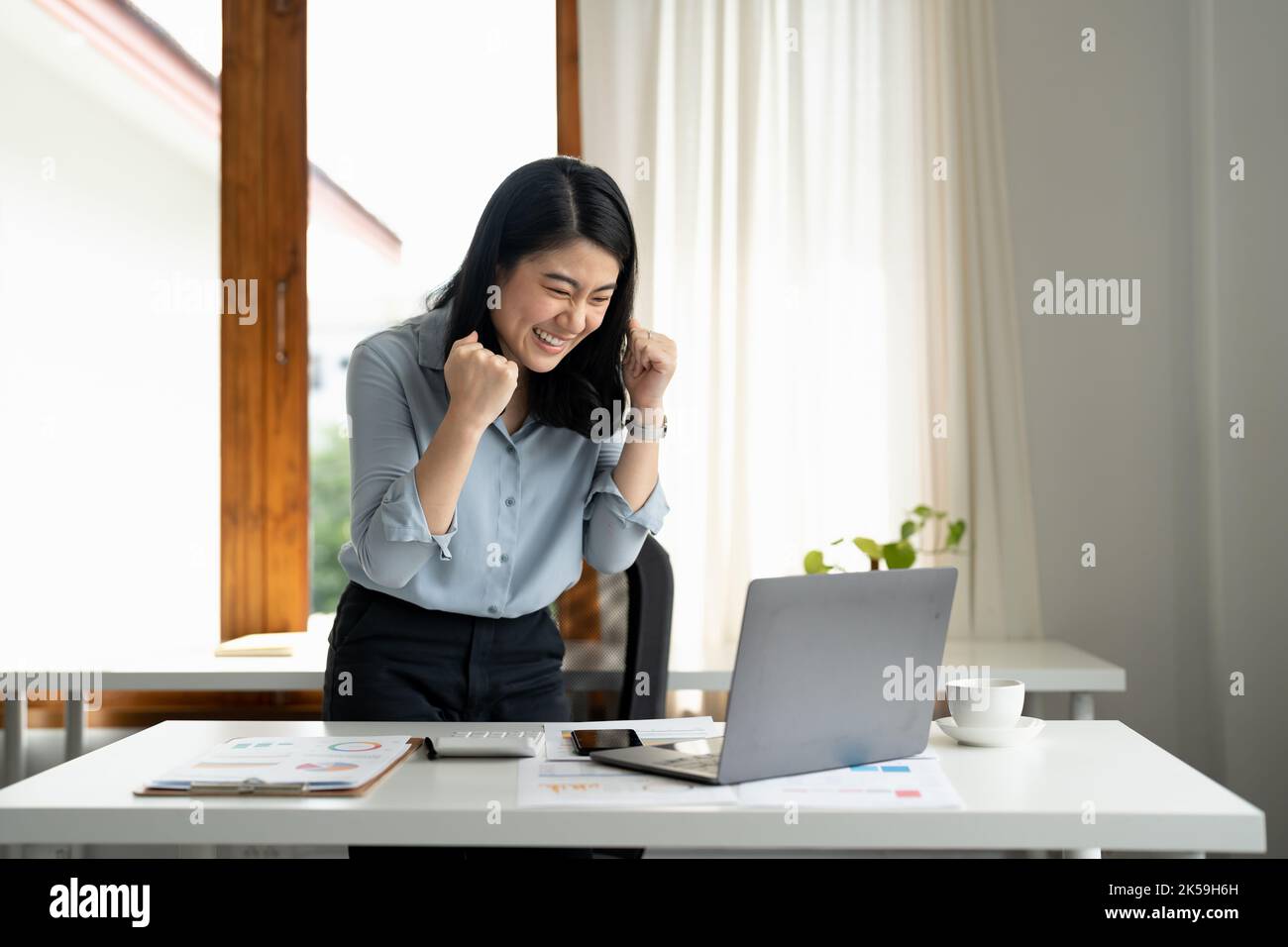 Excited young woman standing at table with laptop and celebrating ...