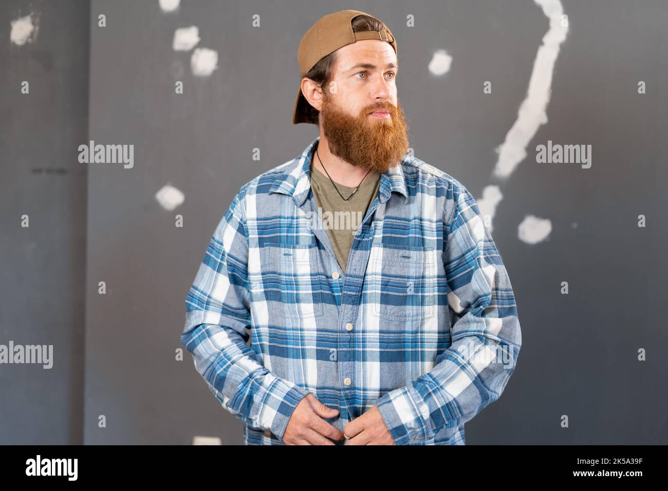 Bearded man in a shirt and a baseball cap on the background of the wall ...