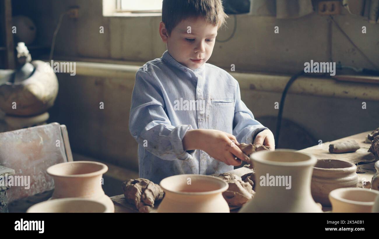 Curious little boy is playing with clay at working table in potter's ...