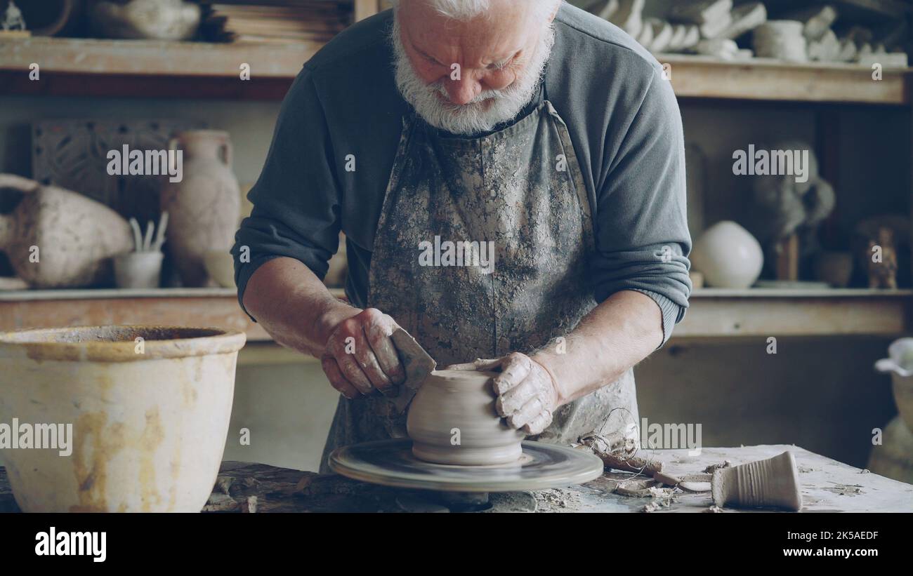 Senior male potter is working at bottom part of ceramic pot on spinning ...