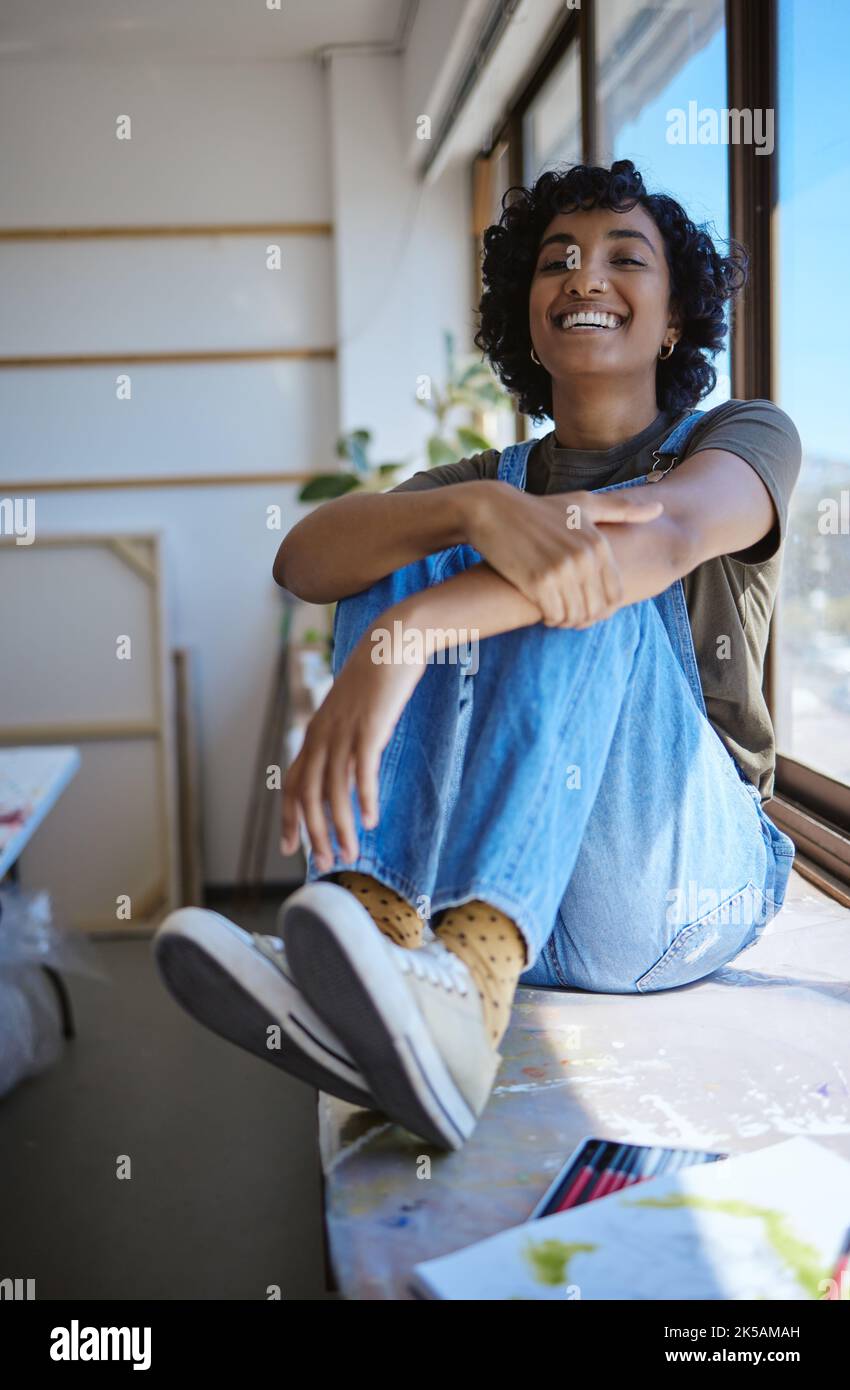 Young woman, artist and by table with window relax, happy and smile in workshop or studio ...