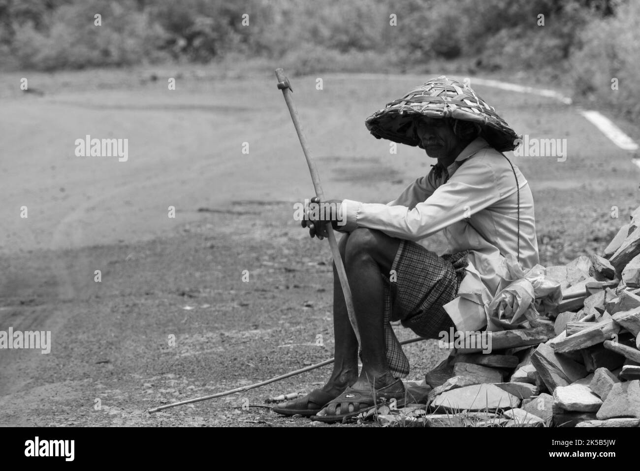A grayscale of an old Indian peasant man resting on rocks on a blurred ...
