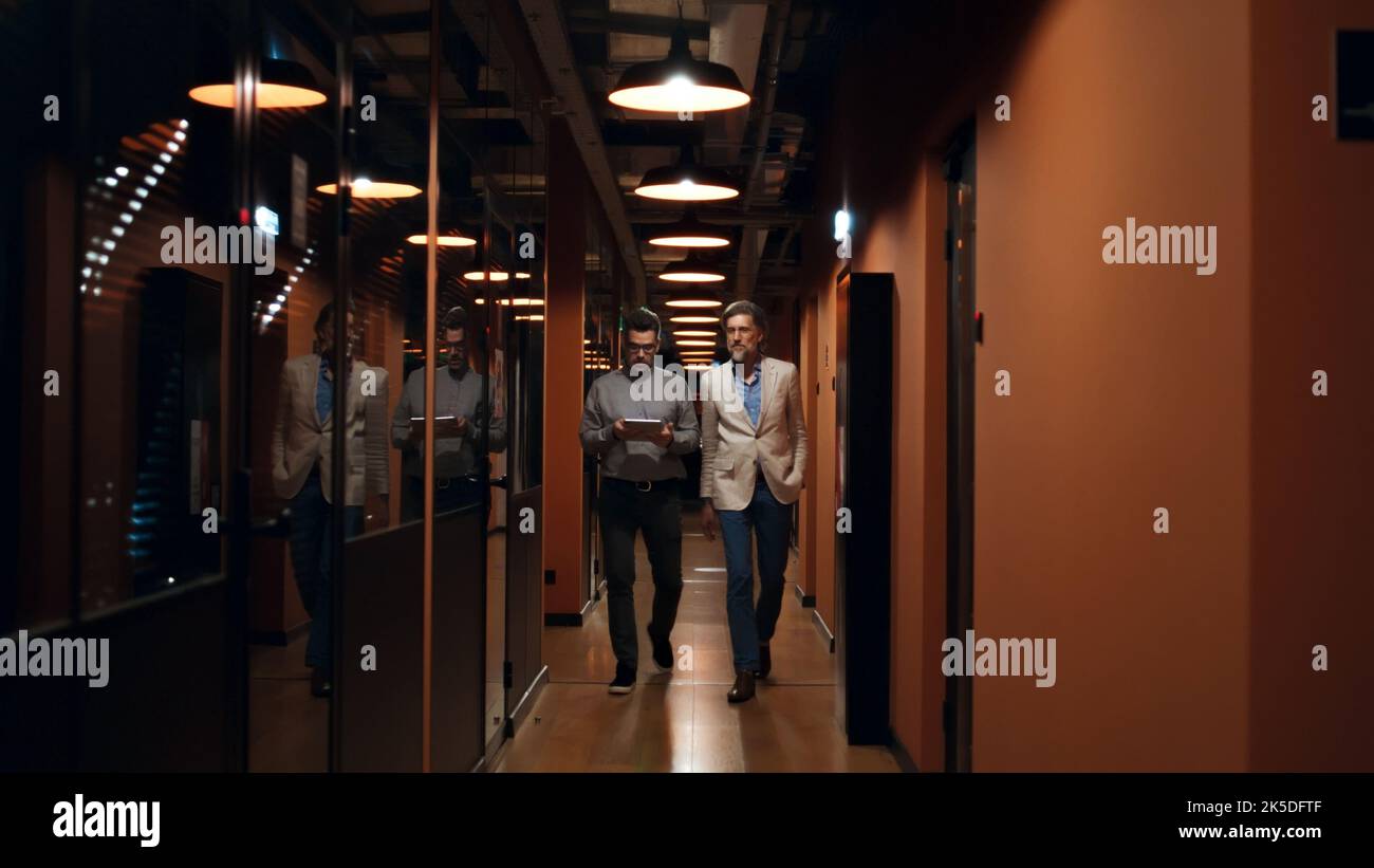 Male coworkers walking down in hallway together, talking about work ...