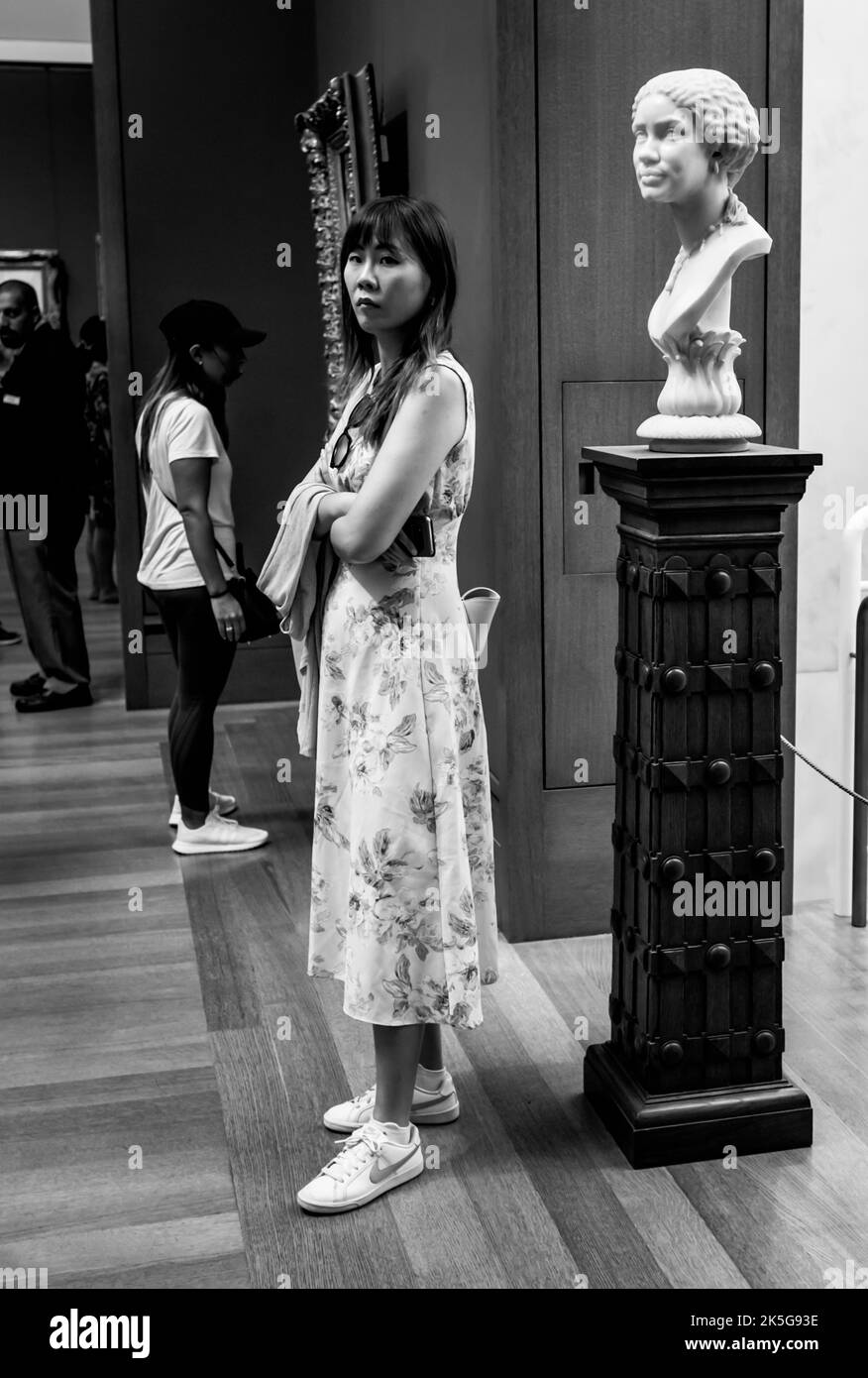A grayscale shot of an Asian lady standing next to a sculpture Inside ...