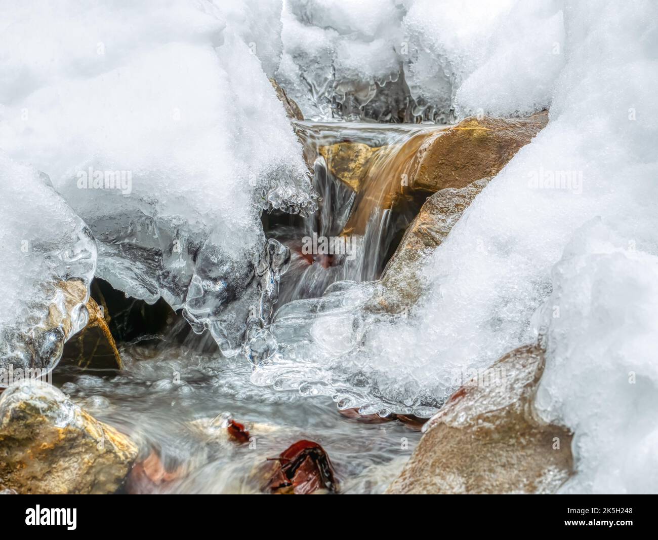 Mountain stream flowing under the white snow through the rocks and ice ...