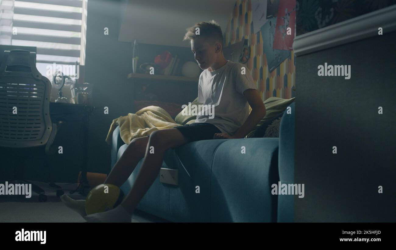 A teenager boy applying bandage to his bruised knee. Sitting on the bed ...