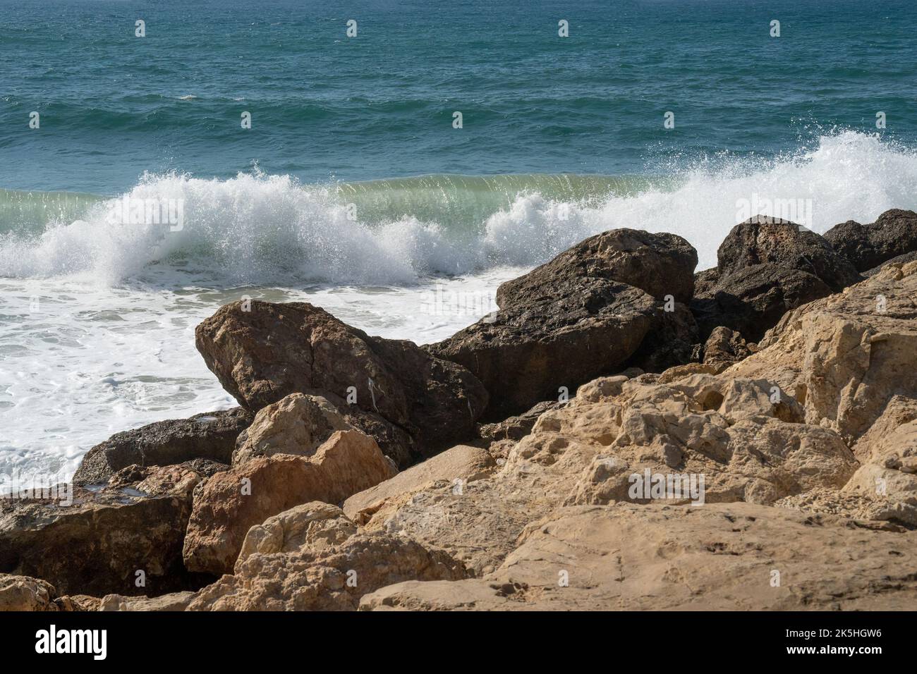 A wave breaking on a rocky shore in Tel Aviv, Israel, on a clear, sunny ...