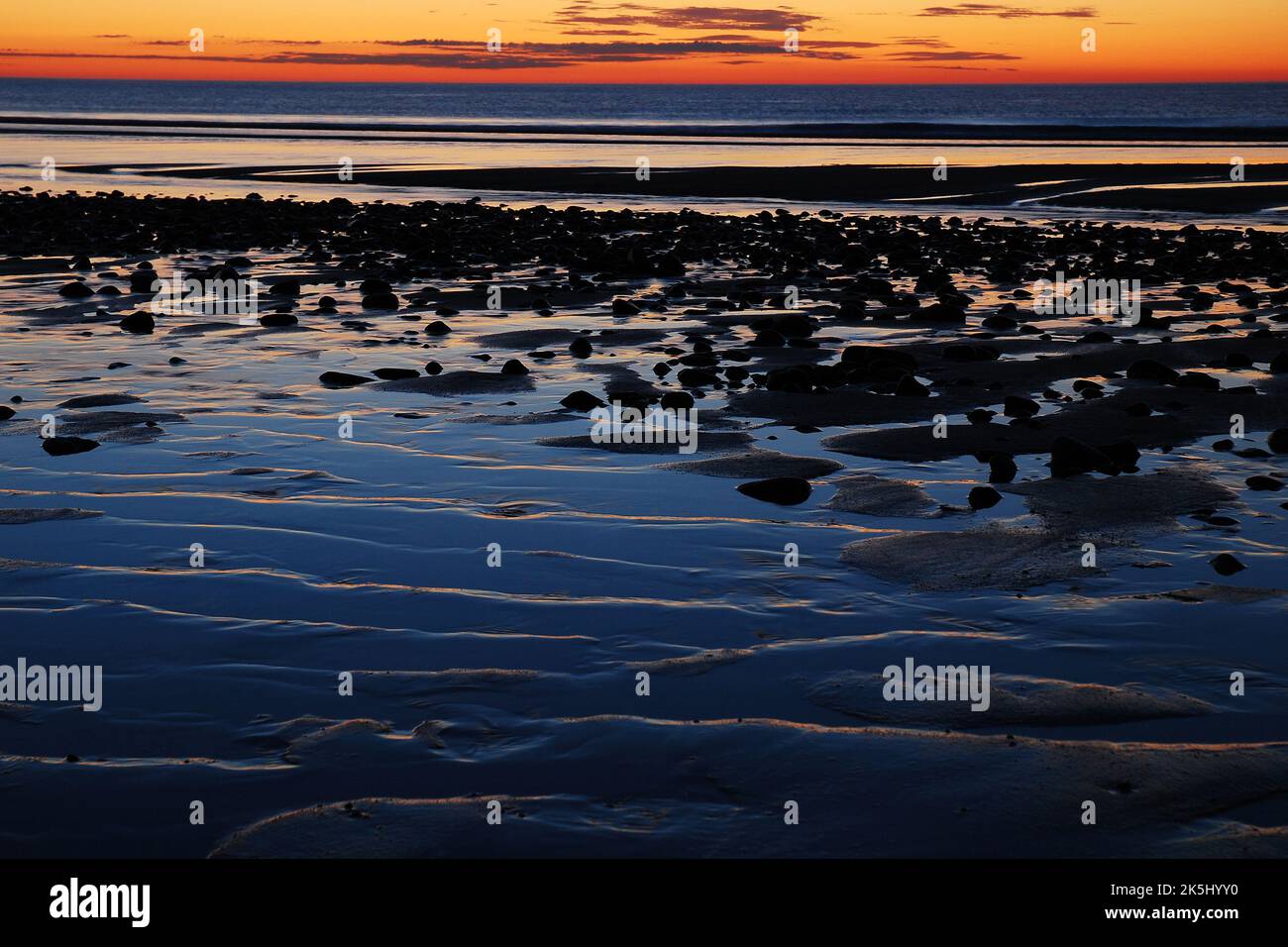 The sunrise sky is reflected in the shallow water and rippled and rocky ...
