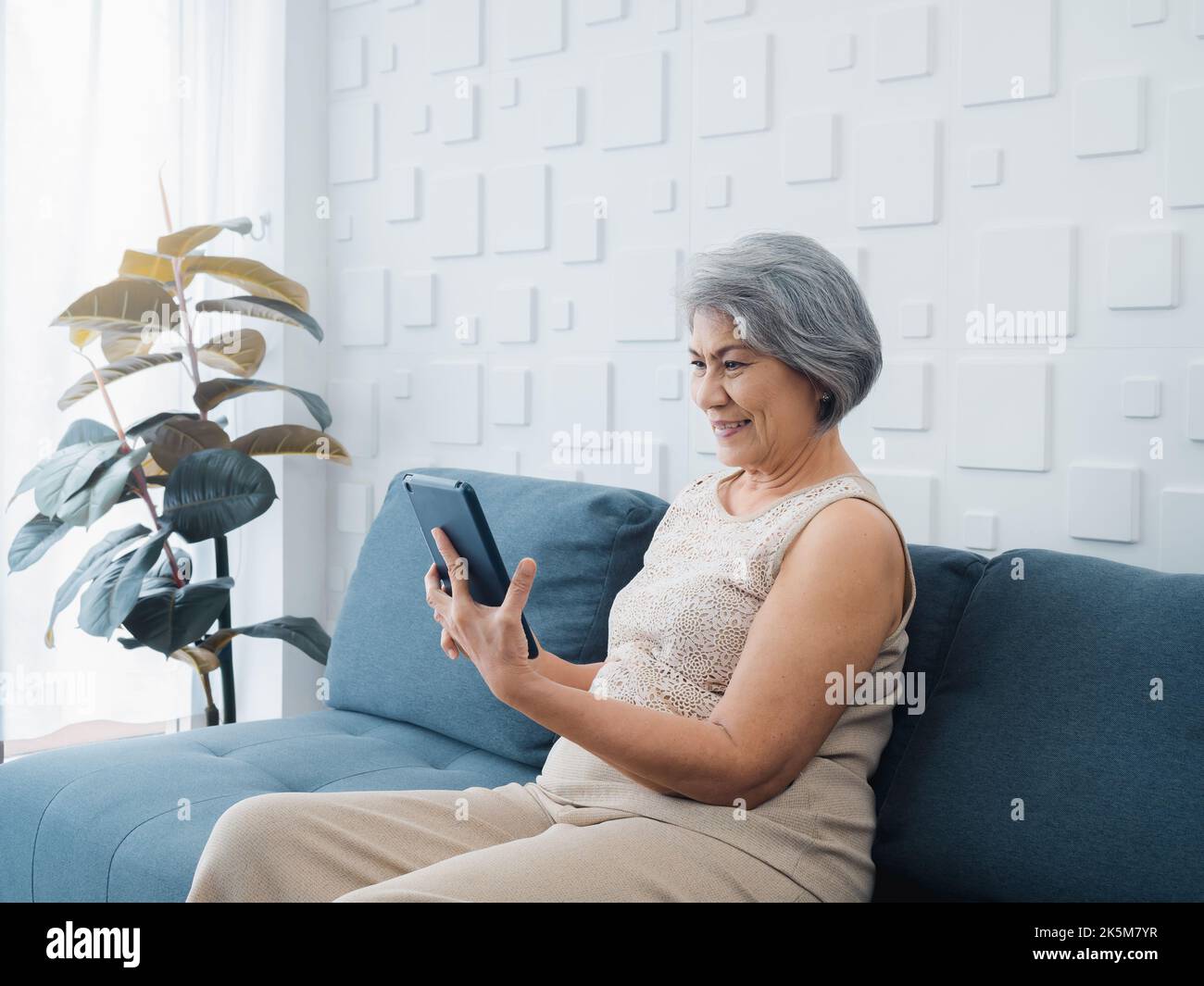 Happy Asian senior woman sit on couch, holding and looking at digital ...