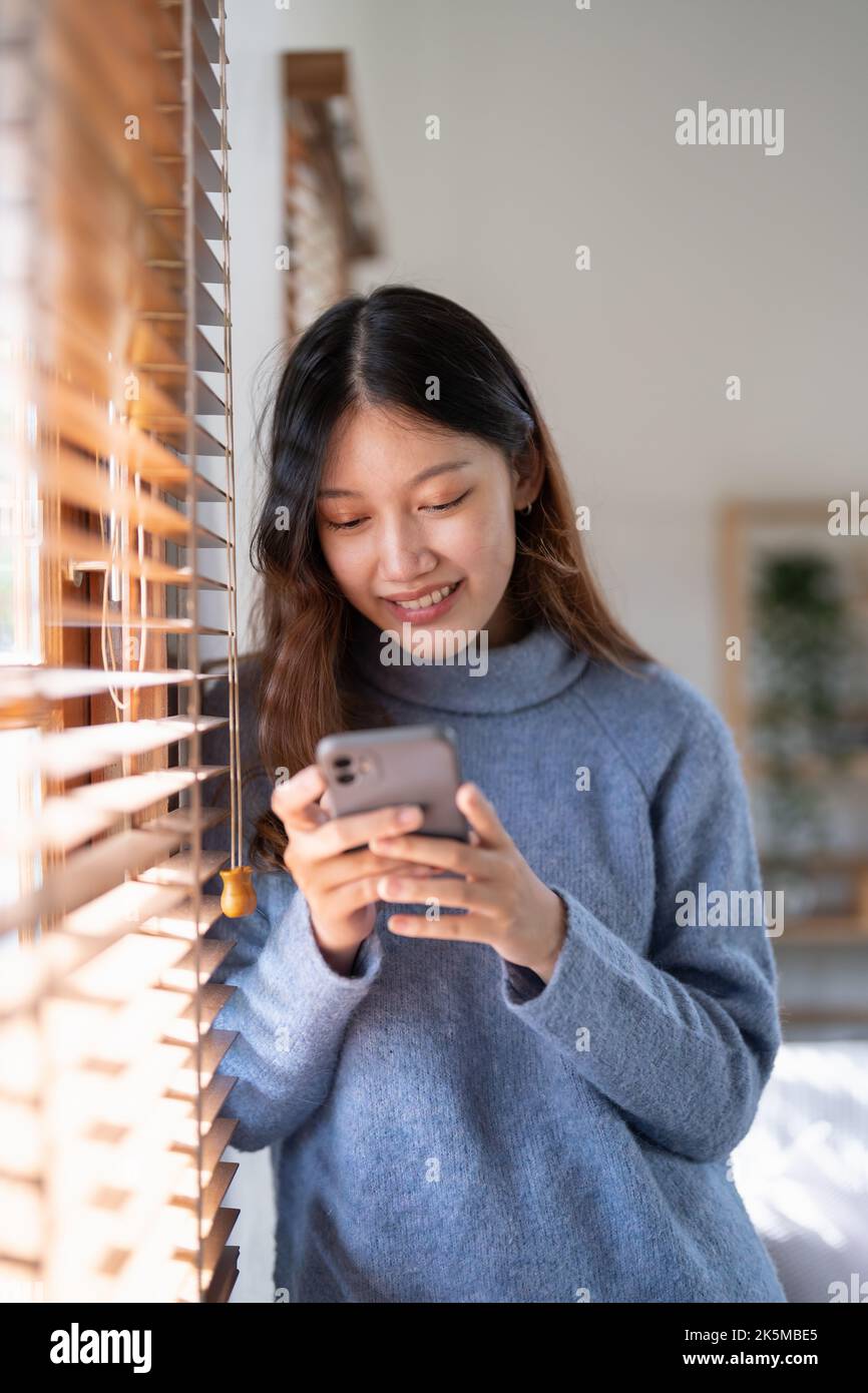 Cheerful young asian woman using mobile phone near window at home Stock ...