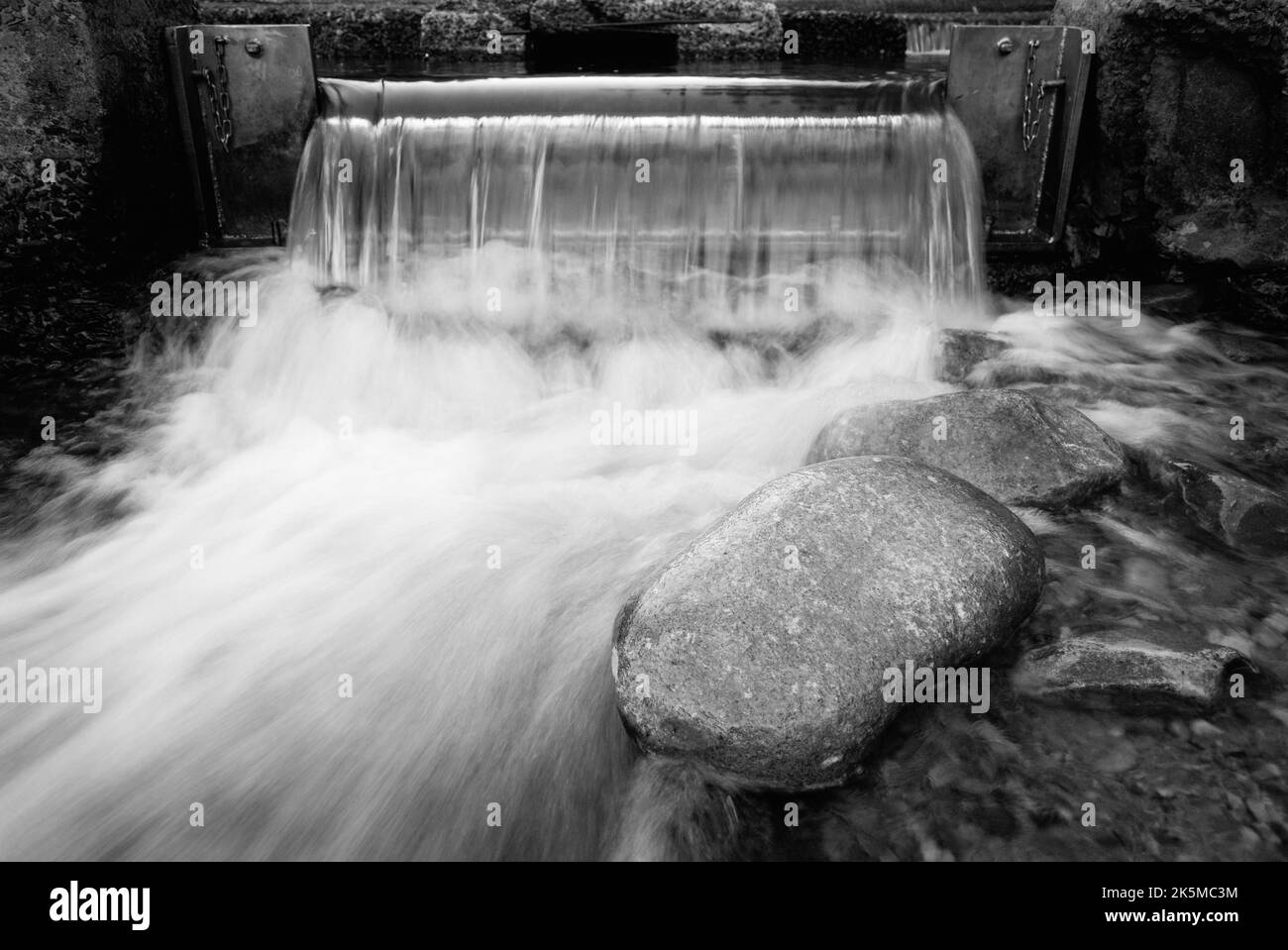 A grayscale of water flowing over weir and hitting the rocks below ...