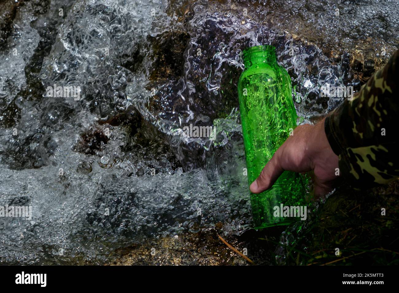 Hand of a traveller holds bottle collecting water from mountain stream ...