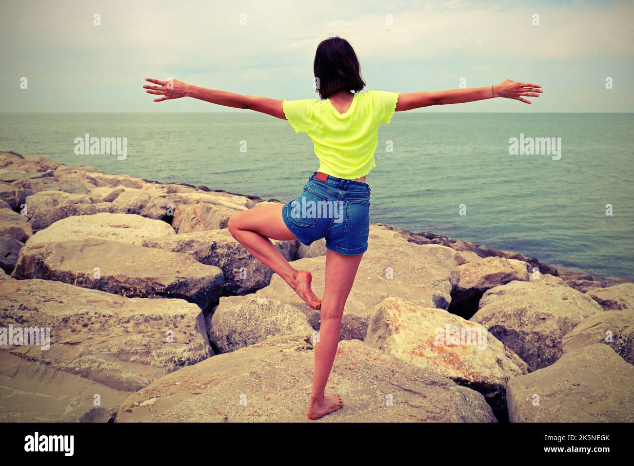 young girl performs gymnastic exercises on the rocks by the sea with ...