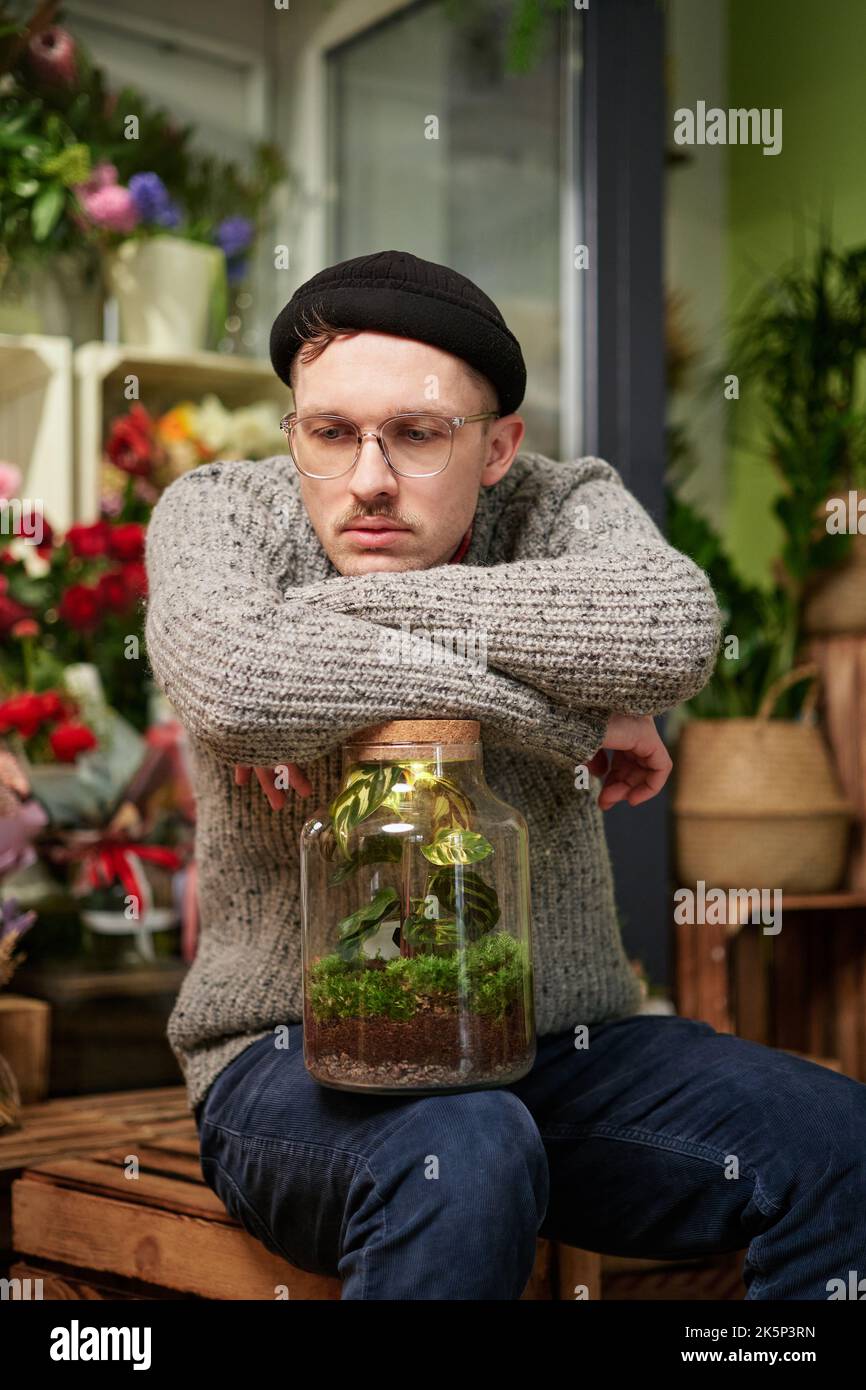 Handsome greenhouse owner or gardener with plants in a jar. Attractive male tired hipster ...