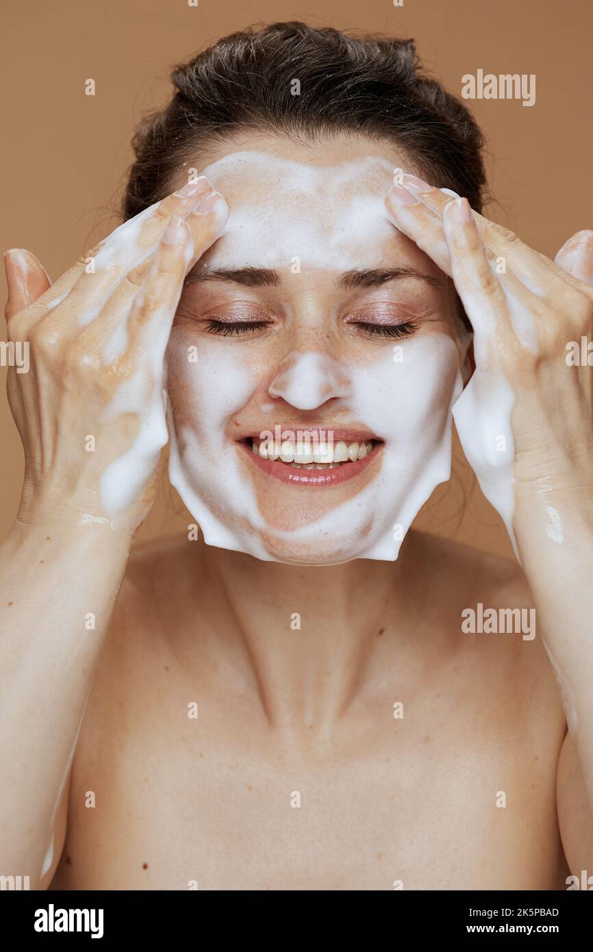 happy middle aged woman with foaming facial cleanser washing face isolated on beige background ...