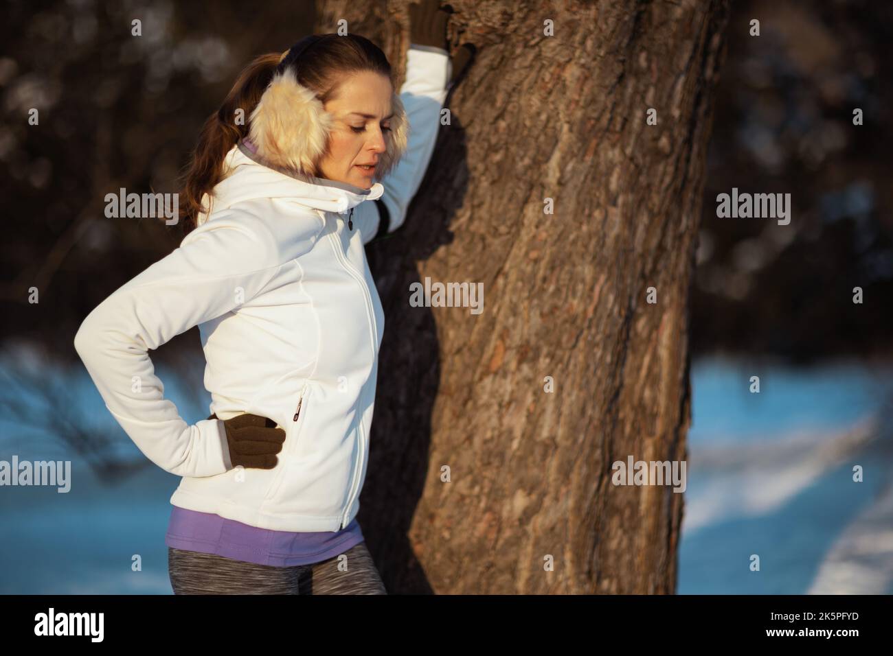 healthy woman in white jacket catching breath after exercise near tree ...