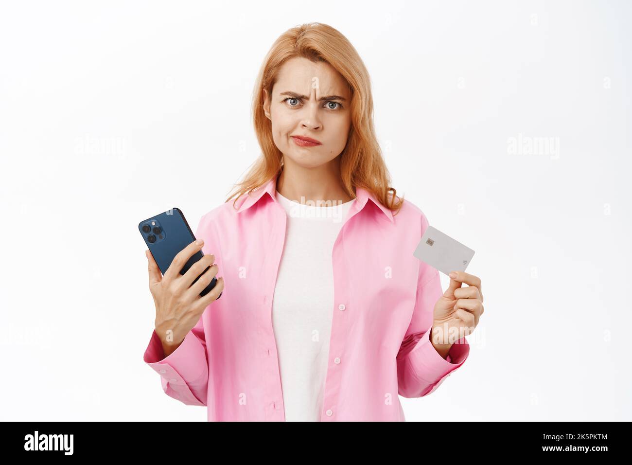 Upset girl with smartphone and credit card, frowns and looks ...