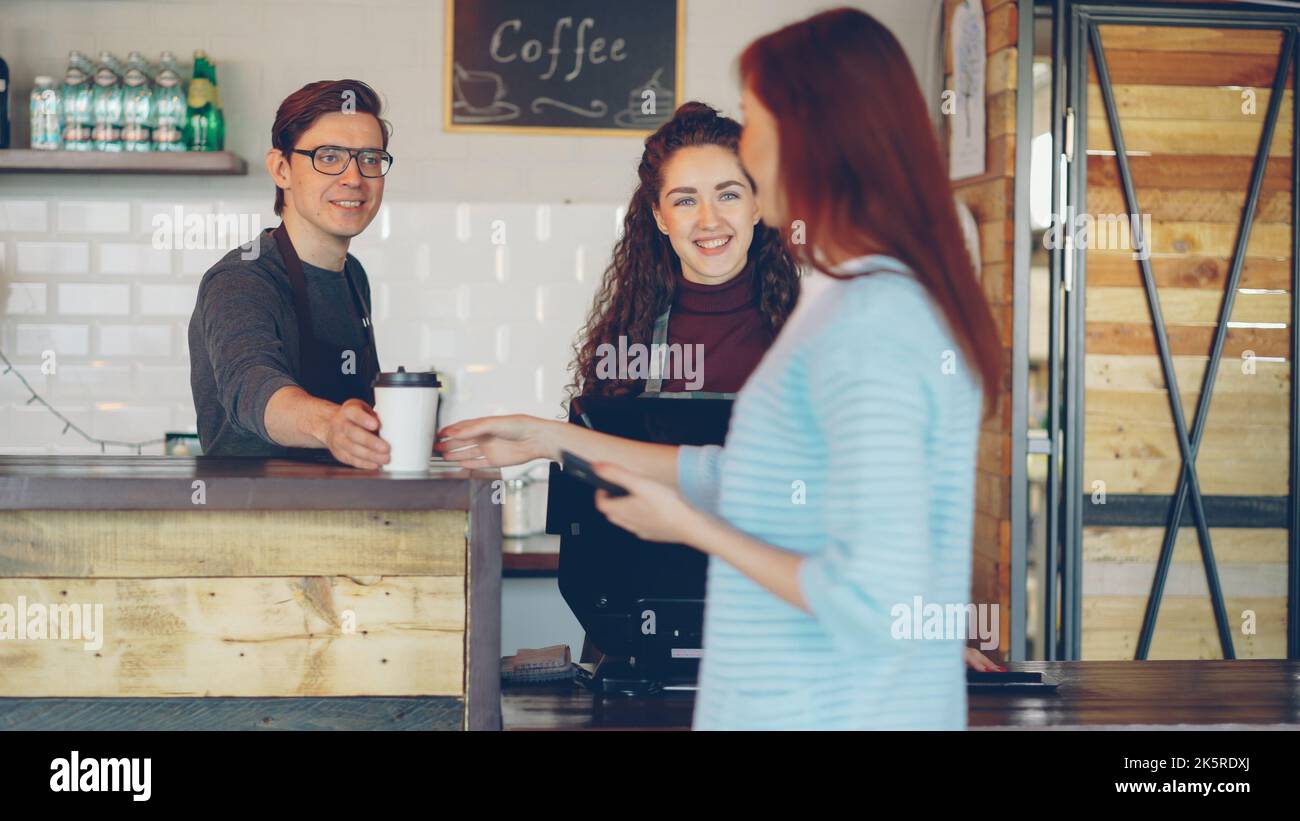 Cheerful female cashier is selling takeaway coffee to young woman and ...