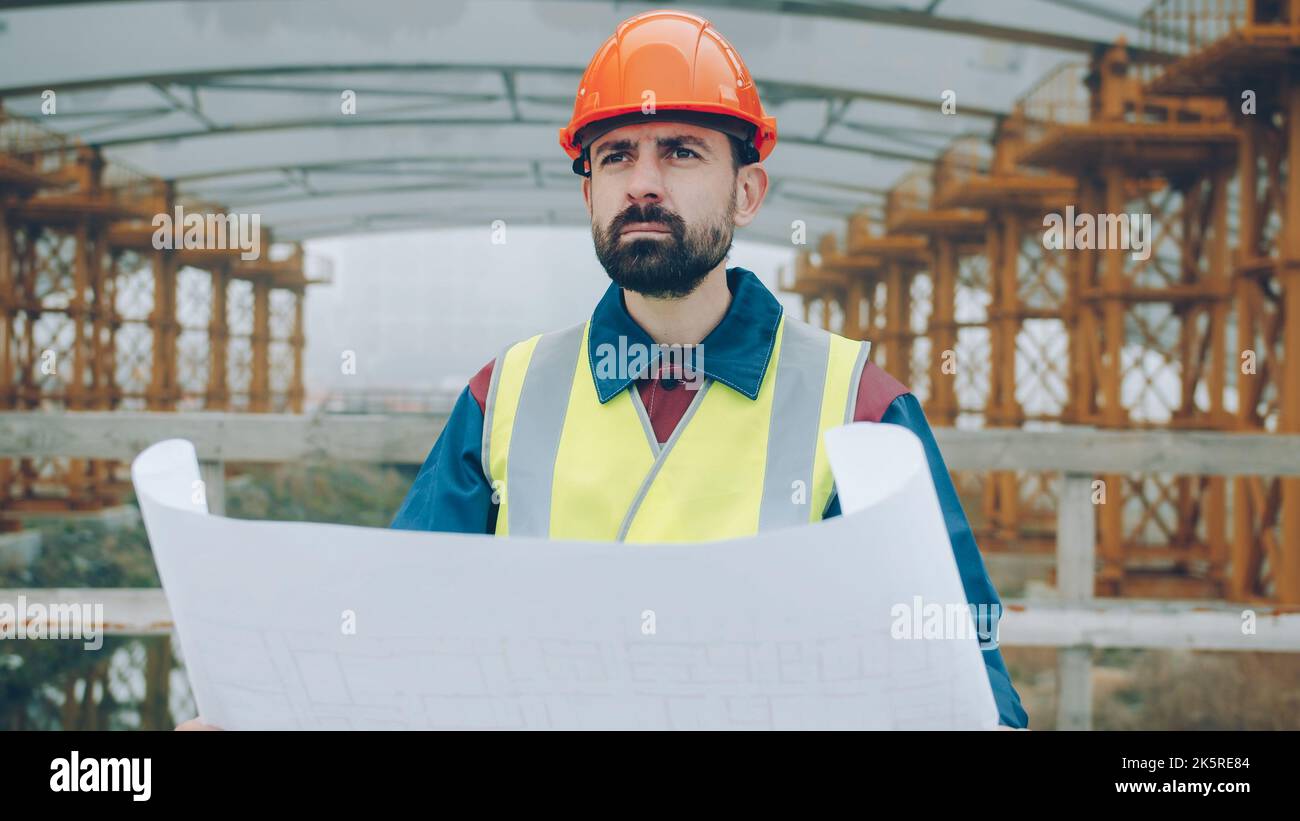 Portrait of serious foreman wearing uniform checking blueprint and ...