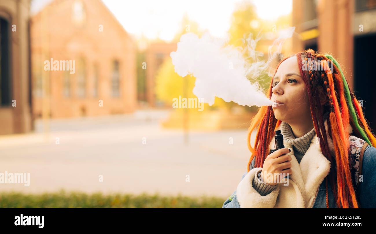Portrait of young woman with dreadlocks vaping, standing on street ...