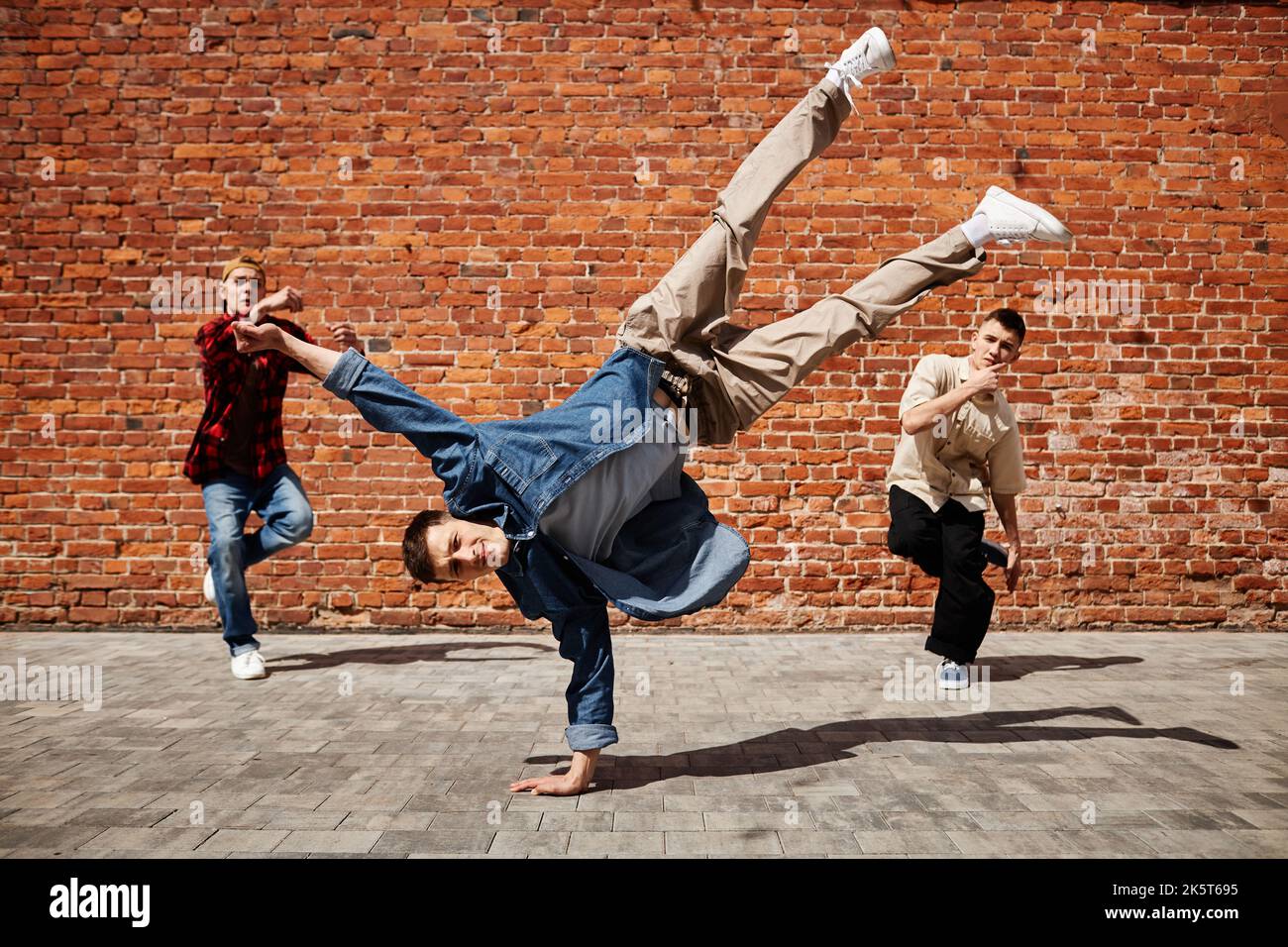 Freeze frame of male breakdance performer doing handstand pose with team against brick wall ...