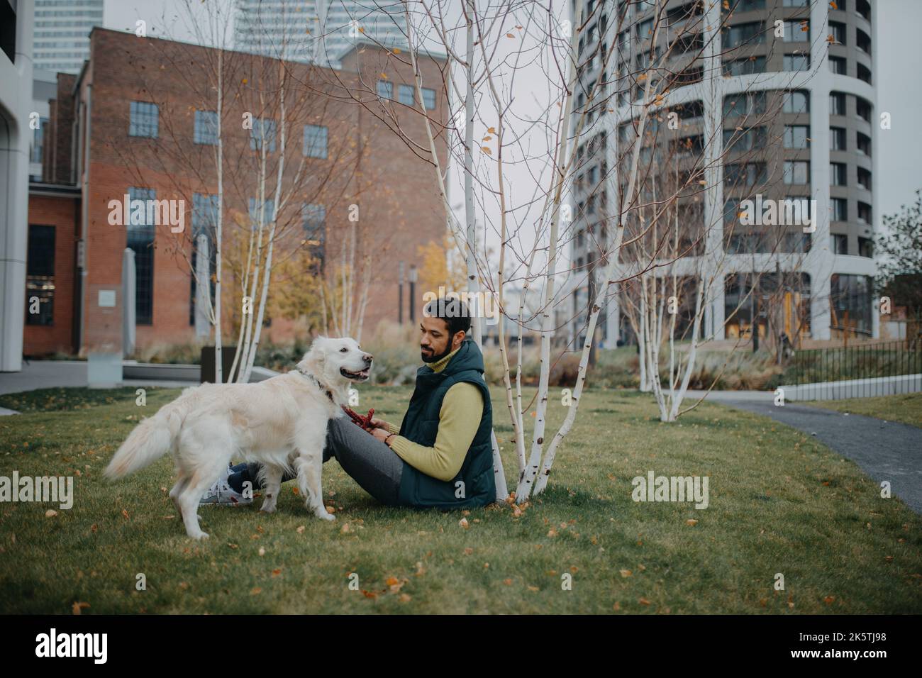 Happy young man stroking his dog outdoors in city park, during cold ...