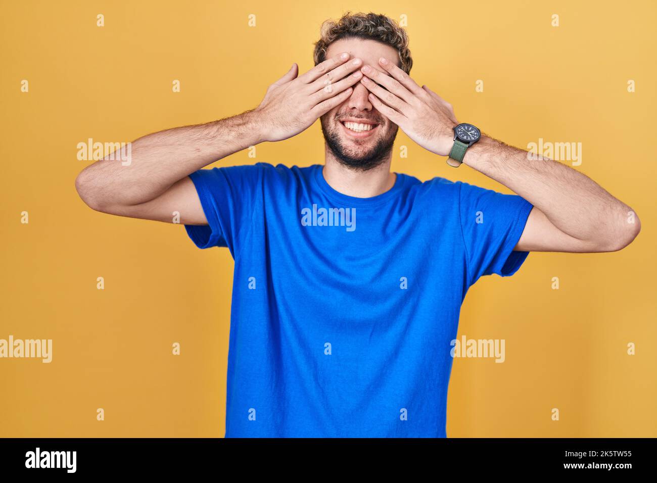 Hispanic man with beard standing over yellow background covering eyes ...