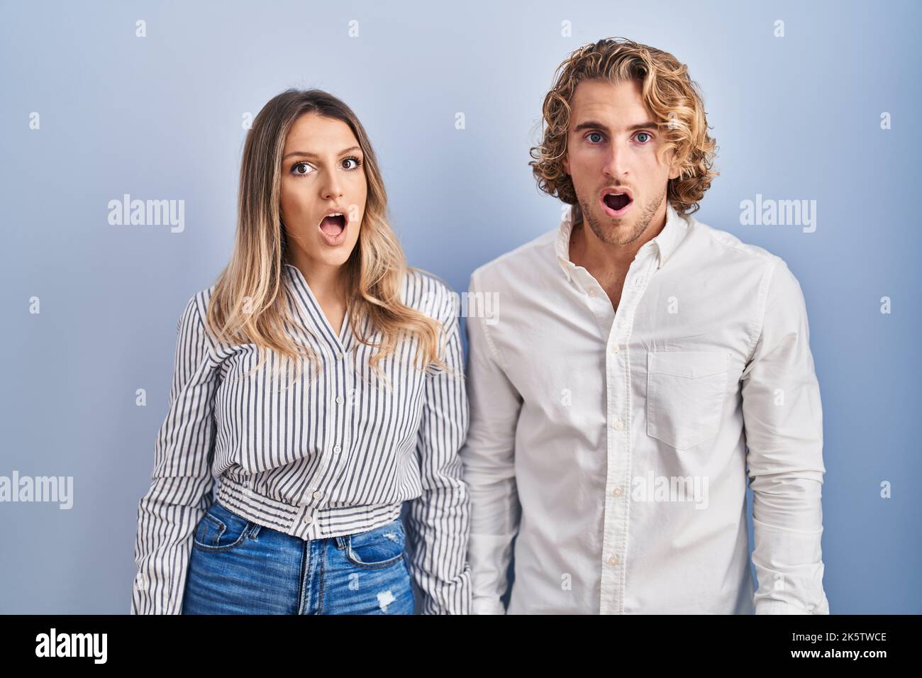 Young couple standing over blue background in shock face, looking ...
