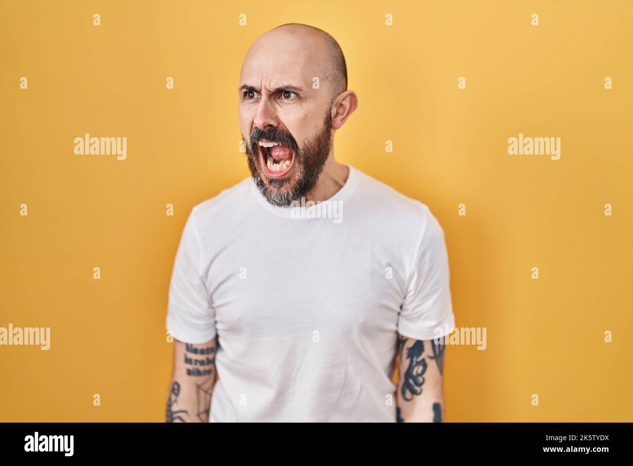 Young hispanic man with tattoos standing over yellow background angry ...