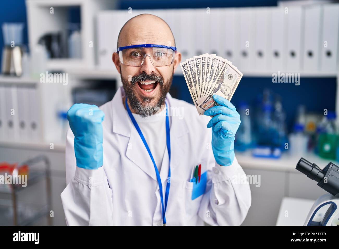 Young hispanic man working at scientist laboratory holding money screaming proud, celebrating ...