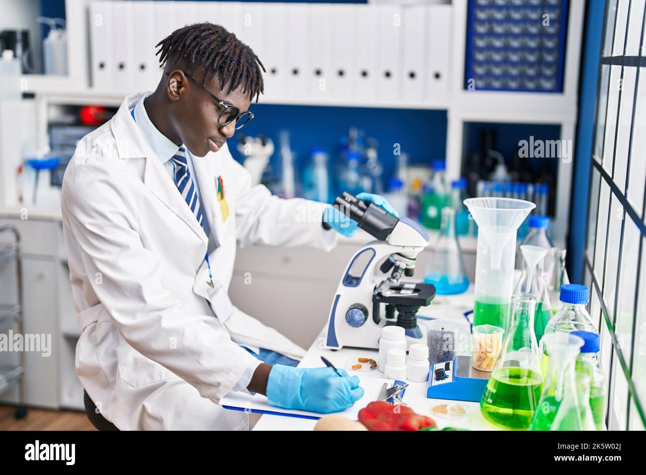 African american man scientist using microscope writing report at laboratory Stock Photo - Alamy